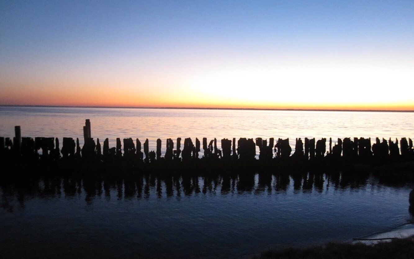 Silhueta de um cais com pessoas no final, refletindo em águas calmas ao pôr do sol.