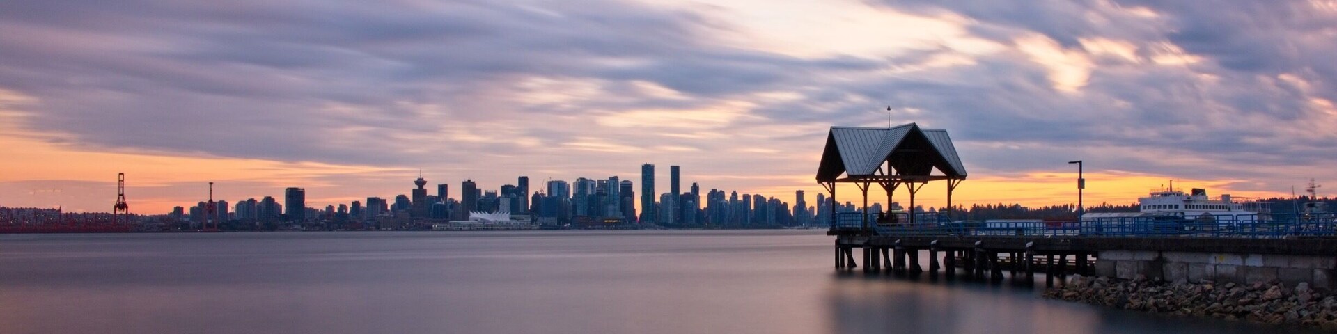 Silhueta urbana ao pôr do sol, com um cais e um coreto.