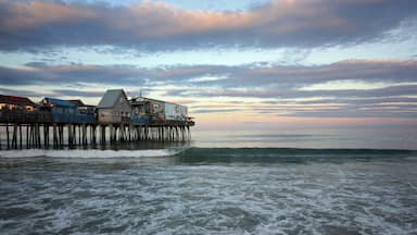 Um cais que se estende para o oceano, com edifícios e ondas quebrando na costa.