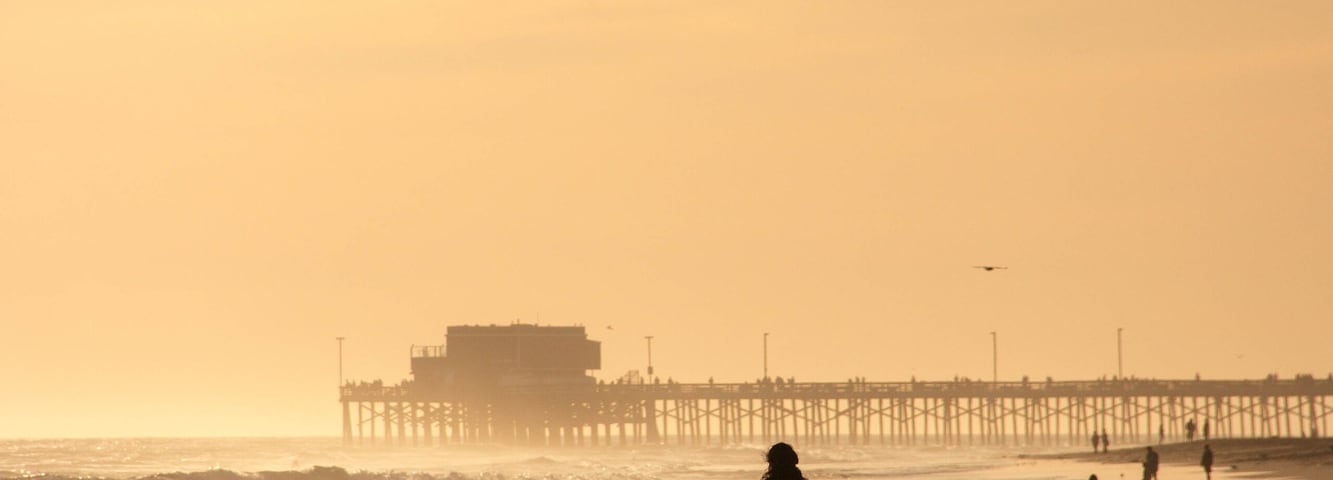 Uma praia com pessoas caminhando e brincando, um píer ao fundo e um céu ao pôr do sol.