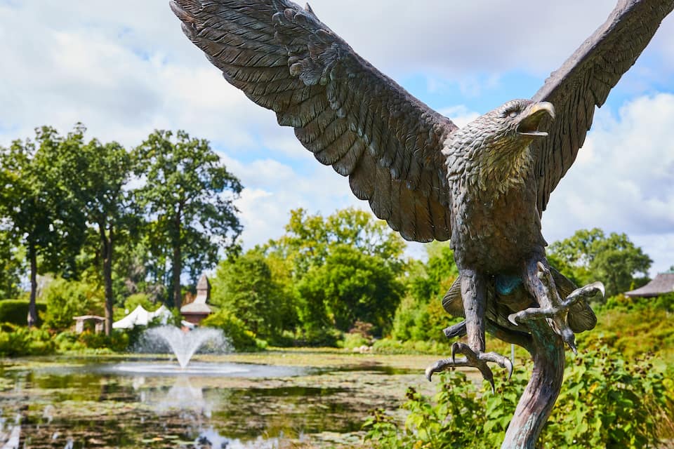 Escultura de bronze de uma águia com asas abertas pousada num galho de árvore, com um lago, vegetação exuberante e uma fonte ao fundo.