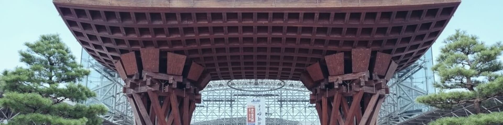 Uma entrada ampla e aberta com telhado de metal e vigas de madeira, rodeada por árvores e uma estrutura de vidro ao fundo.