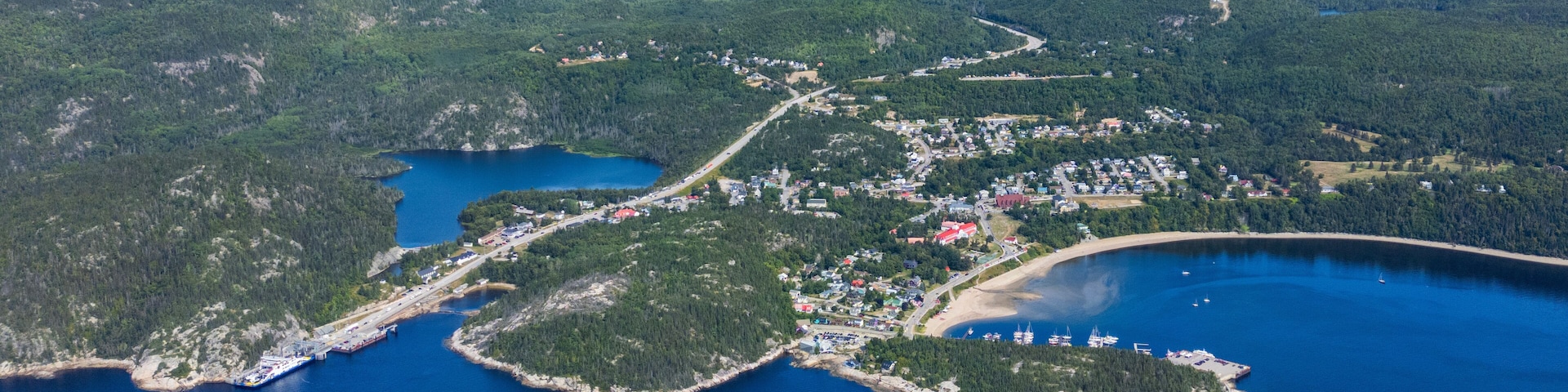 Vista aérea de uma cidade costeira com um porto de iates, rodeada por florestas densas e uma grande extensão de água.