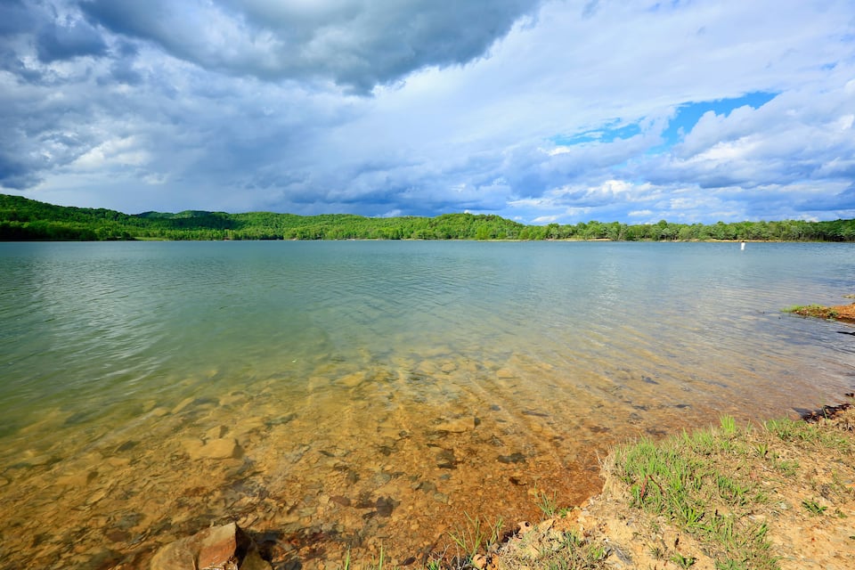 Um lago calmo com águas cristalinas, rodeado por colinas verdes e um céu nublado.
