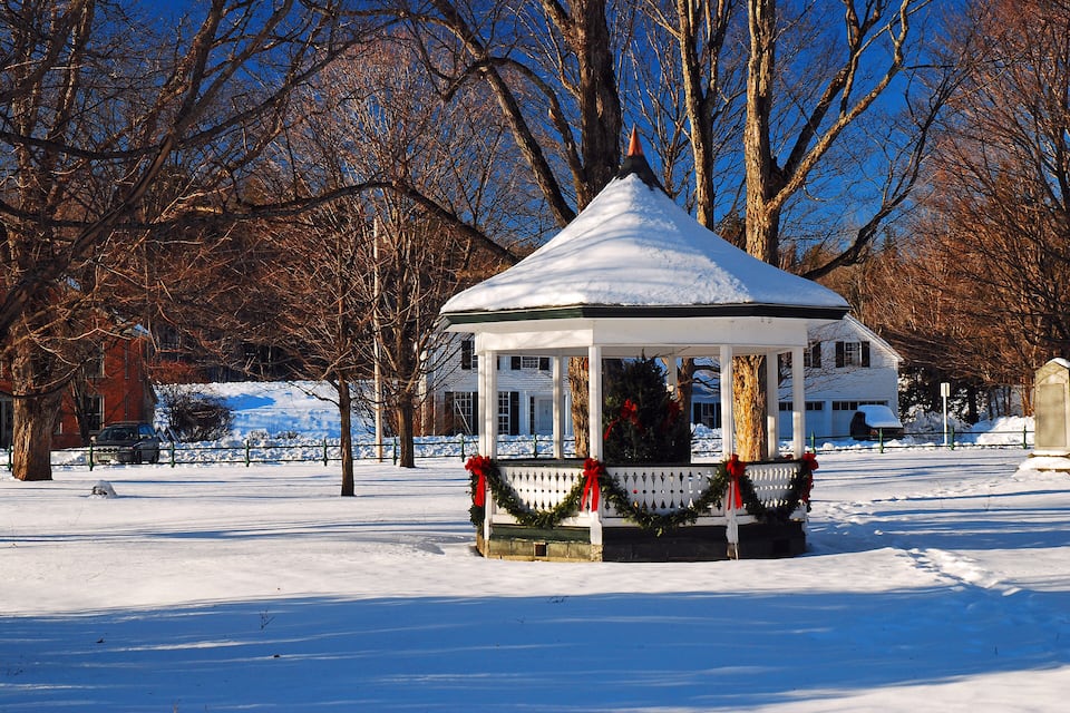 Um gazebo coberto de neve, decorado com laços vermelhos e uma guirlanda, em meio a árvores sem folhas e uma paisagem nevada.