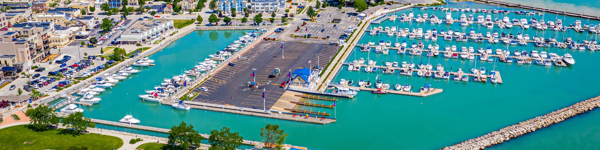 Uma marina com vários barcos atracados, um cais e uma praia com um farol ao fundo.