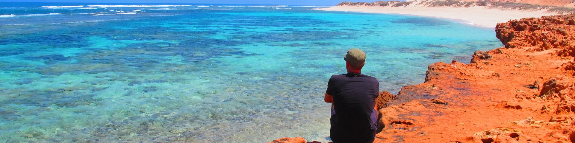 Um homem sentado sobre uma rocha vermelha, com vista para um mar azul límpido e uma praia arenosa.