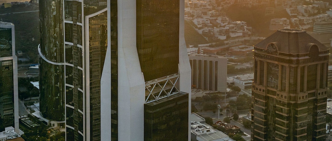 Paisagem urbana com arranha-céus modernos, uma torre imponente com design único e um pôr do sol ao fundo.