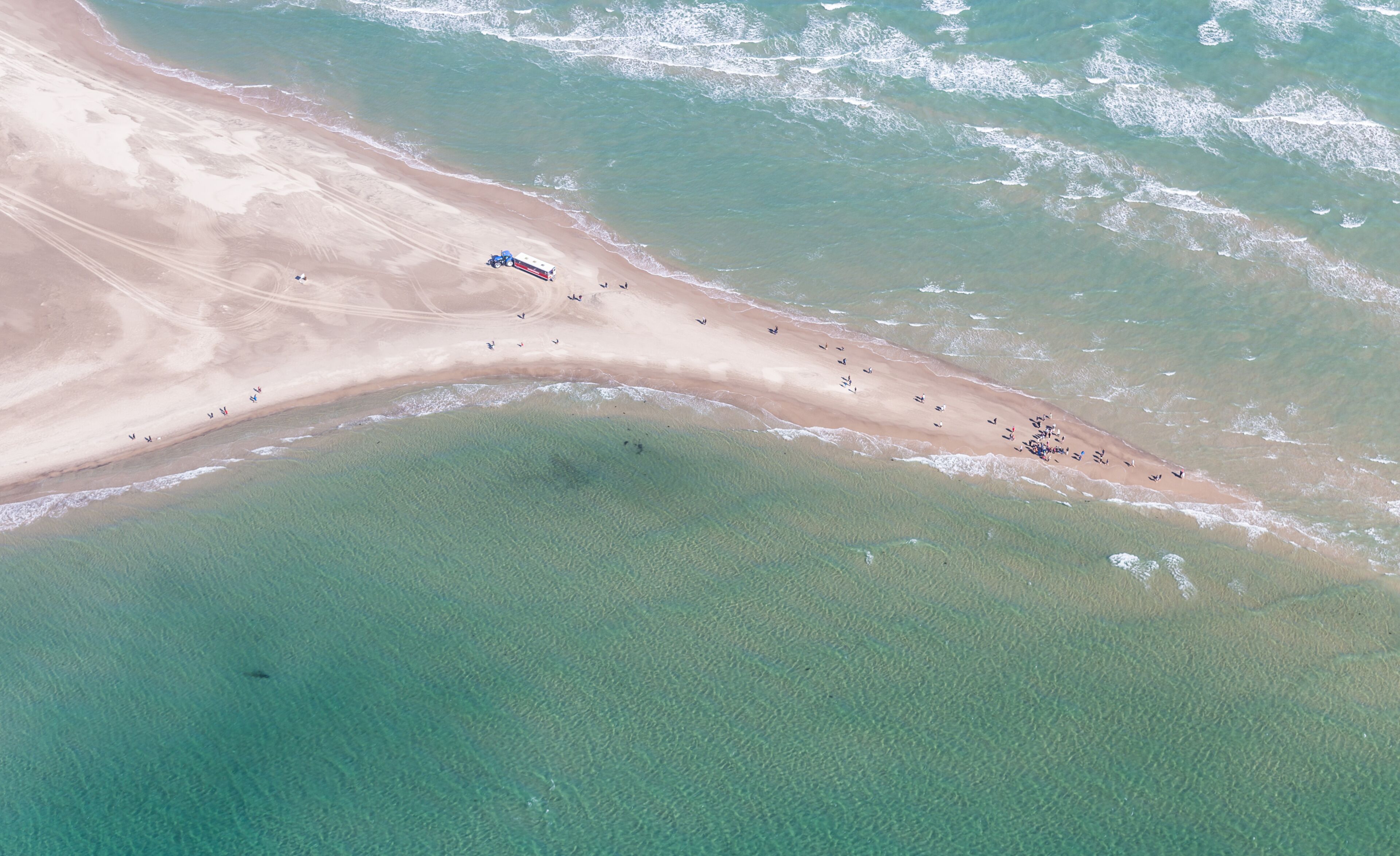 Vista aérea de uma praia com águas turquesa cristalinas, areal arenoso e um ônibus estacionado na areia.