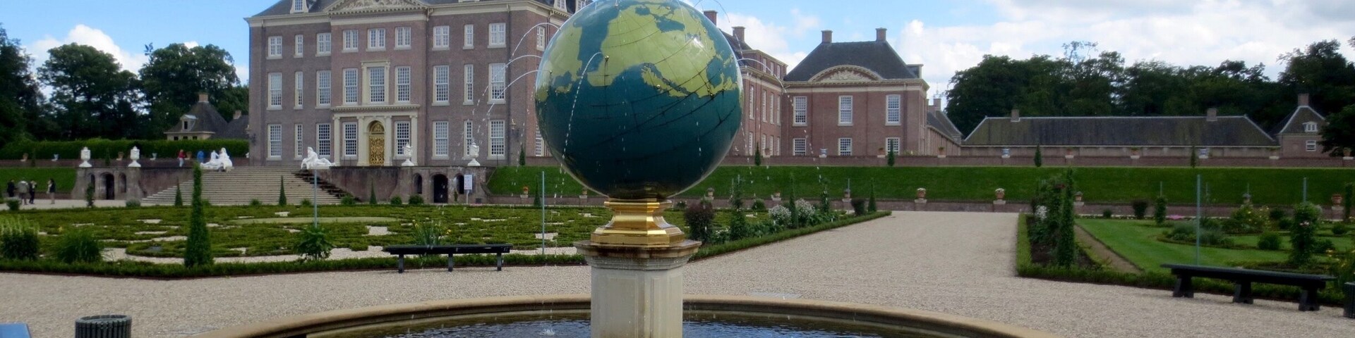 Uma fonte em forma de globo num jardim formal, com um edifício histórico imponente ao fundo.