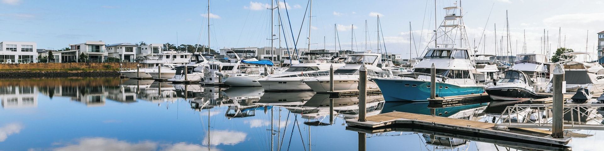 Uma marina com vários barcos atracados, superfície de água calma refletindo o céu e os barcos, e edifícios modernos ao fundo.