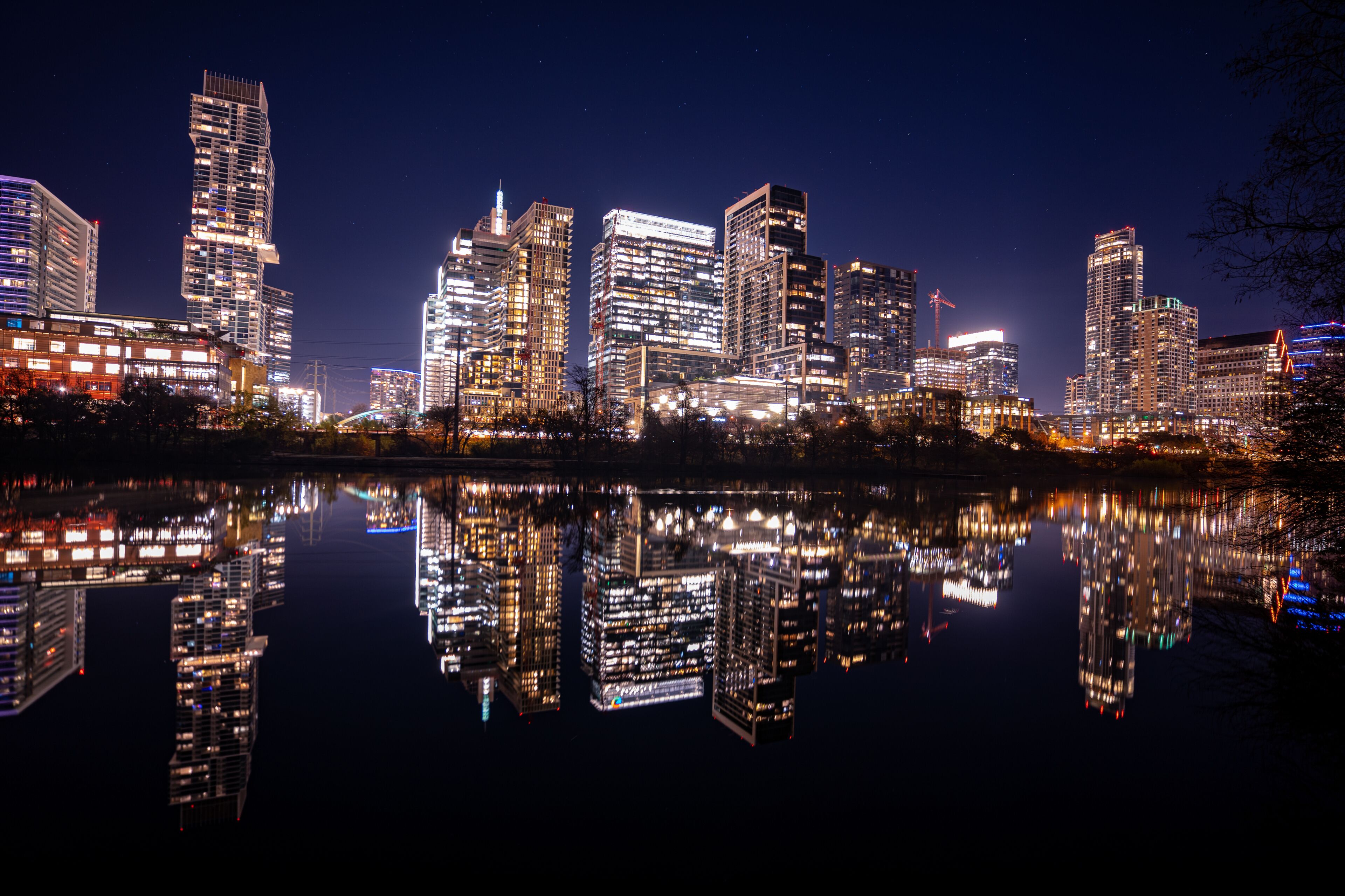 Horizonte urbano noturno com edifícios iluminados refletidos em águas calmas.