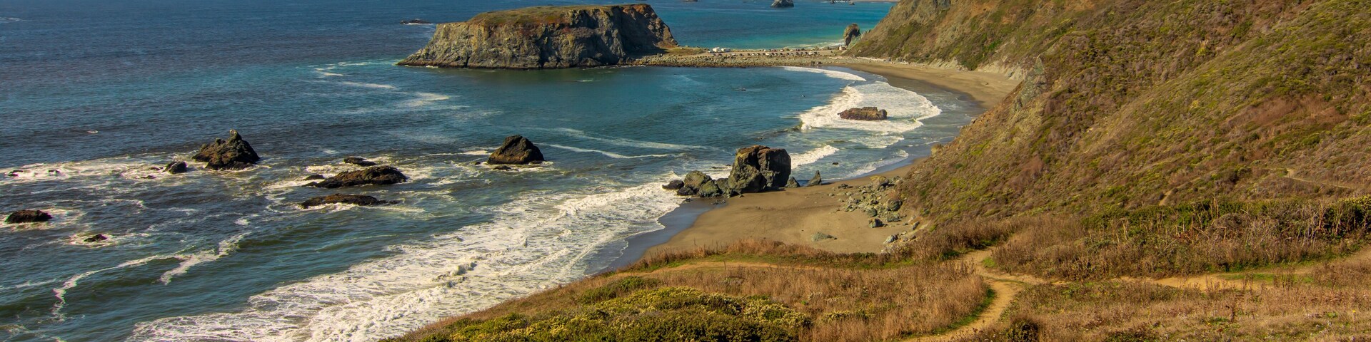 Paisagem costeira com praia arenosa, afloramentos rochosos e céu azul claro.