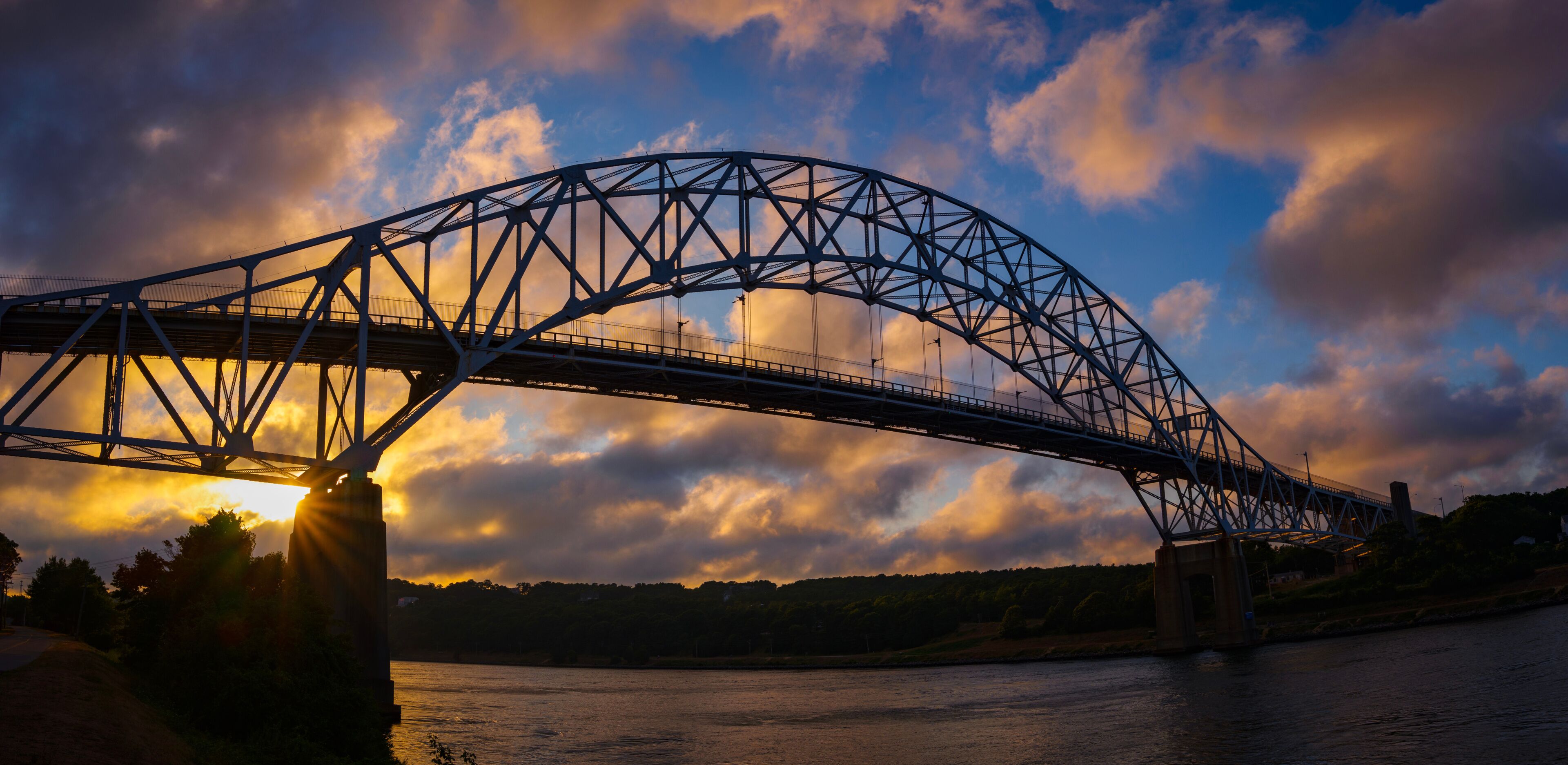 Uma ponte treliçada de aço sobre um rio, com o pôr do sol ao fundo.