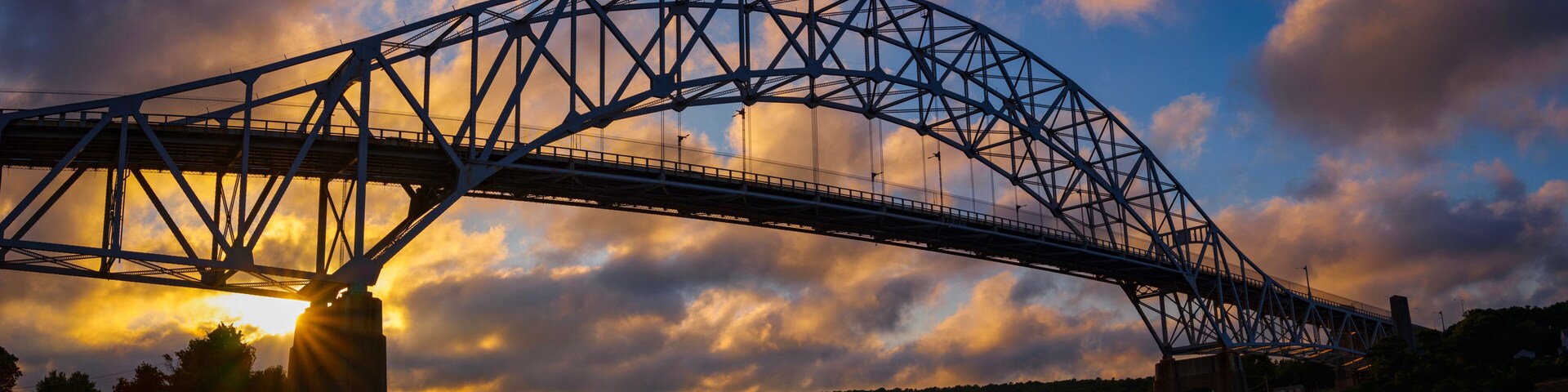 Uma ponte treliçada de aço sobre um rio, com o pôr do sol ao fundo.