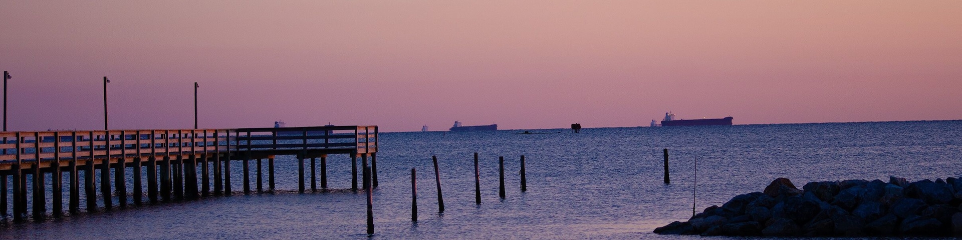 Um píer se estendendo sobre águas calmas ao pôr do sol, com um navio distante.