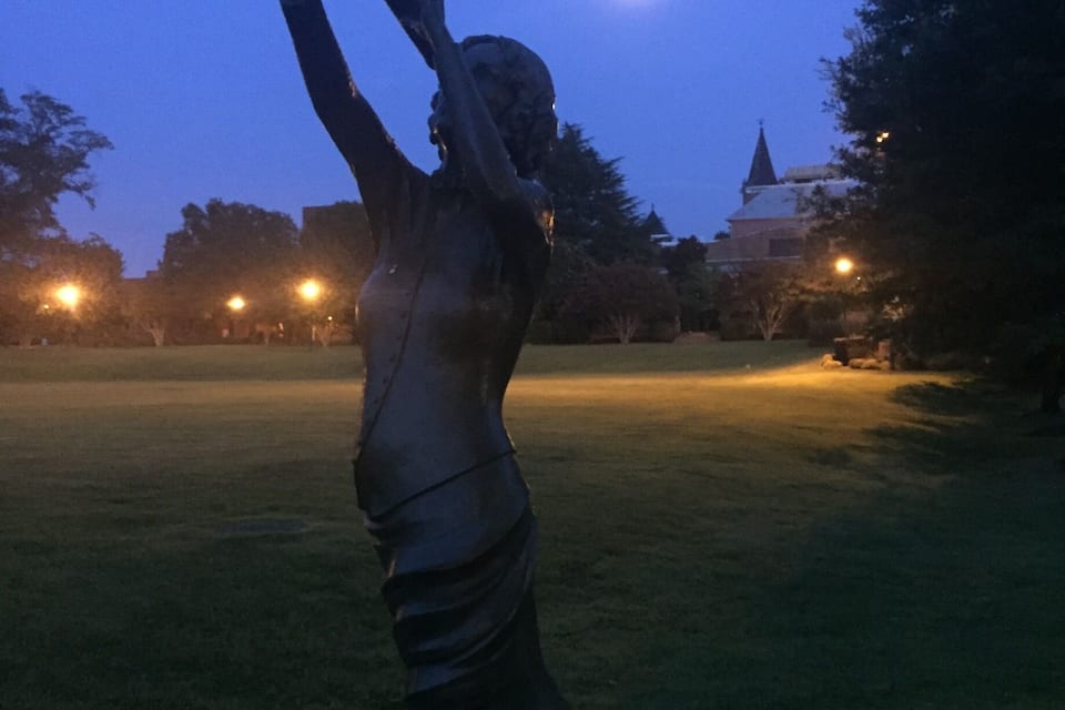 Estátua de uma figura segurando um bastão em um parque ao entardecer.