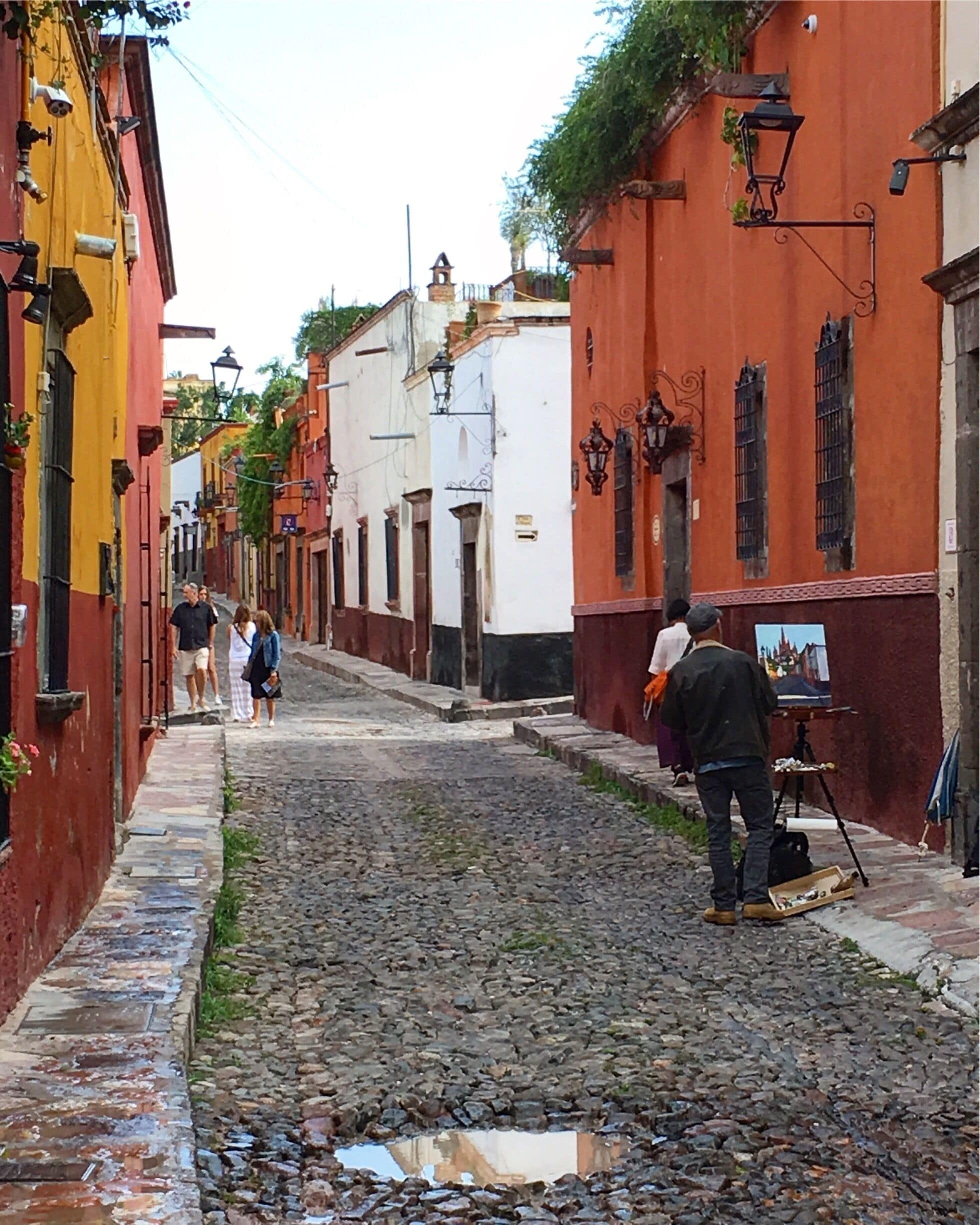 Rua de paralelepípedos ladeada por edifícios coloridos, uma pessoa pintando e uma pequena poça refletindo o ambiente.