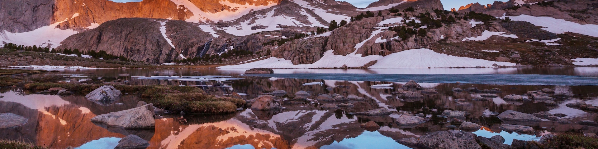 Paisagem de montanha com um lago refletindo os picos, rodeada por manchas de neve e terreno rochoso.