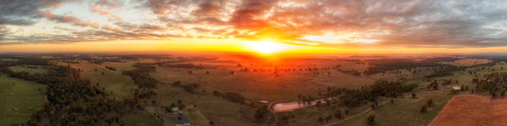 Um pôr do sol sobre um vale com campos, um pequeno corpo d'água e árvores dispersas.