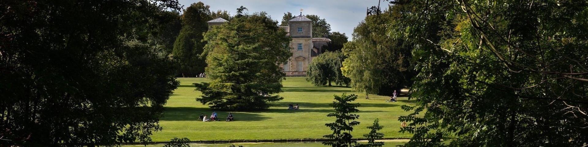Um parque com um gramado extenso, um lago e um edifício com uma torre ao longe.